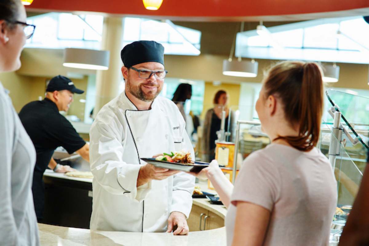 White male cafeteria worker in a chef’s coat serving a female customer